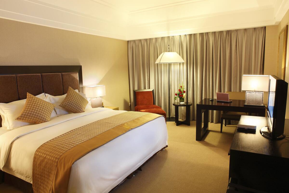 Quarto de hotel com uma cama grande, uma escrivaninha com computador, uma cadeira e uma luminária.