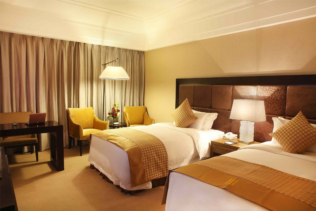 Quarto de hotel com duas camas, uma escrivaninha, uma cadeira e um abajur.