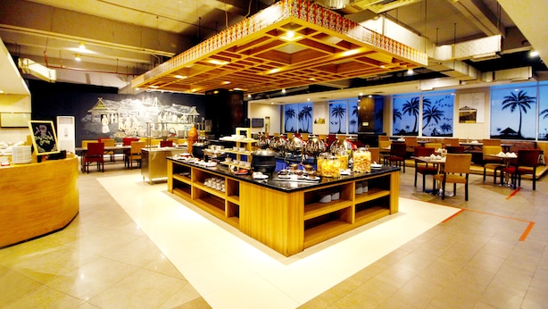 Uma área de buffet com ilha central, diversos arranjos de assentos e teto alto com vigas aparentes.