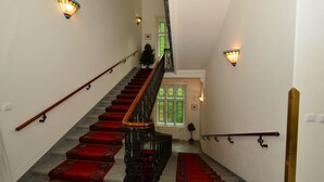 Uma escadaria com degraus revestidos de carpete vermelho, corrimãos de madeira e luminárias fixadas na parede.