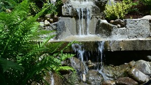 Uma cascata caindo sobre rochas em um jardim com samambaias e uma parede de pedra.