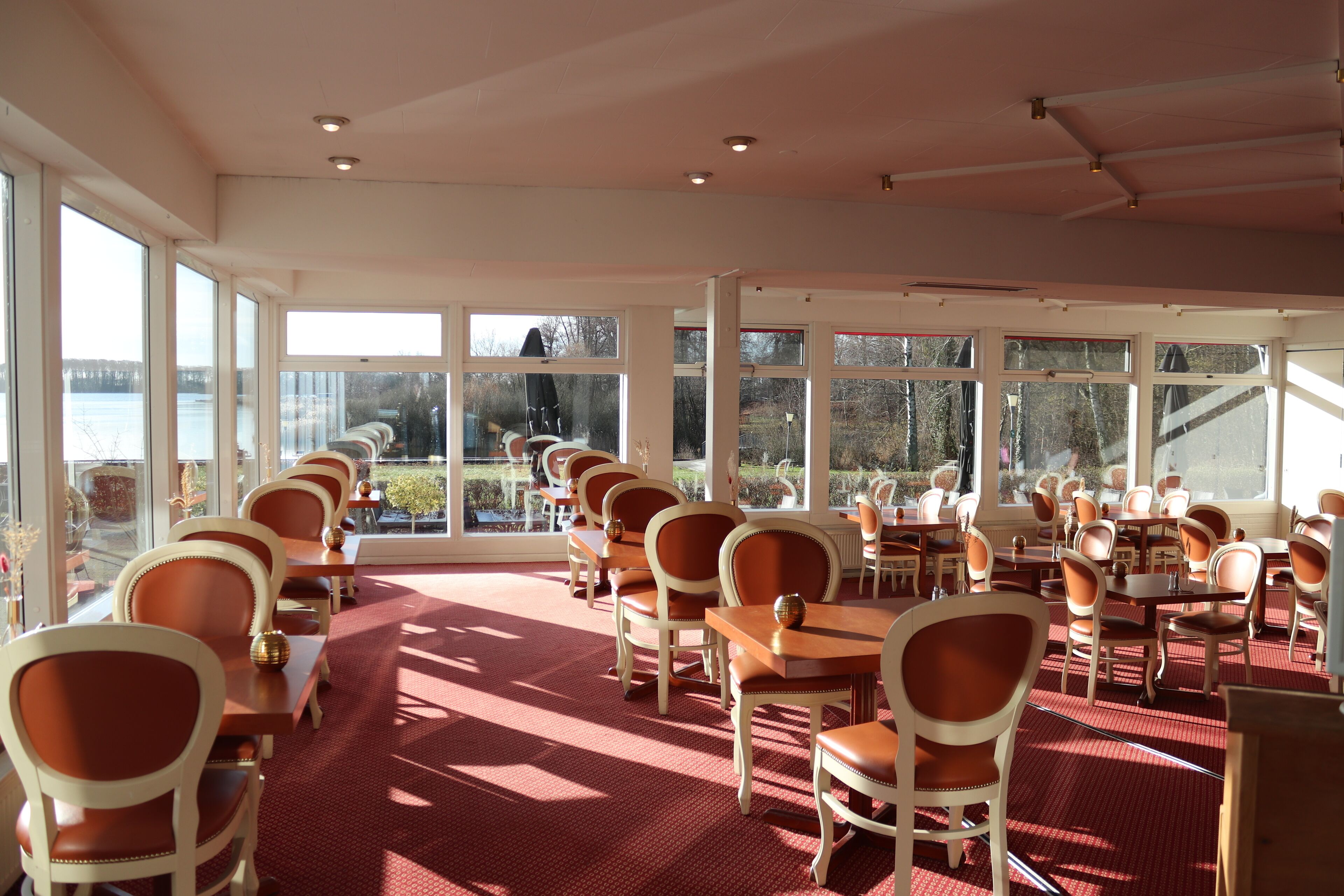 Uma sala de jantar com mesas e cadeiras dispostas para os hóspedes, grandes janelas com vista para o exterior e piso coberto por um tapete vermelho.