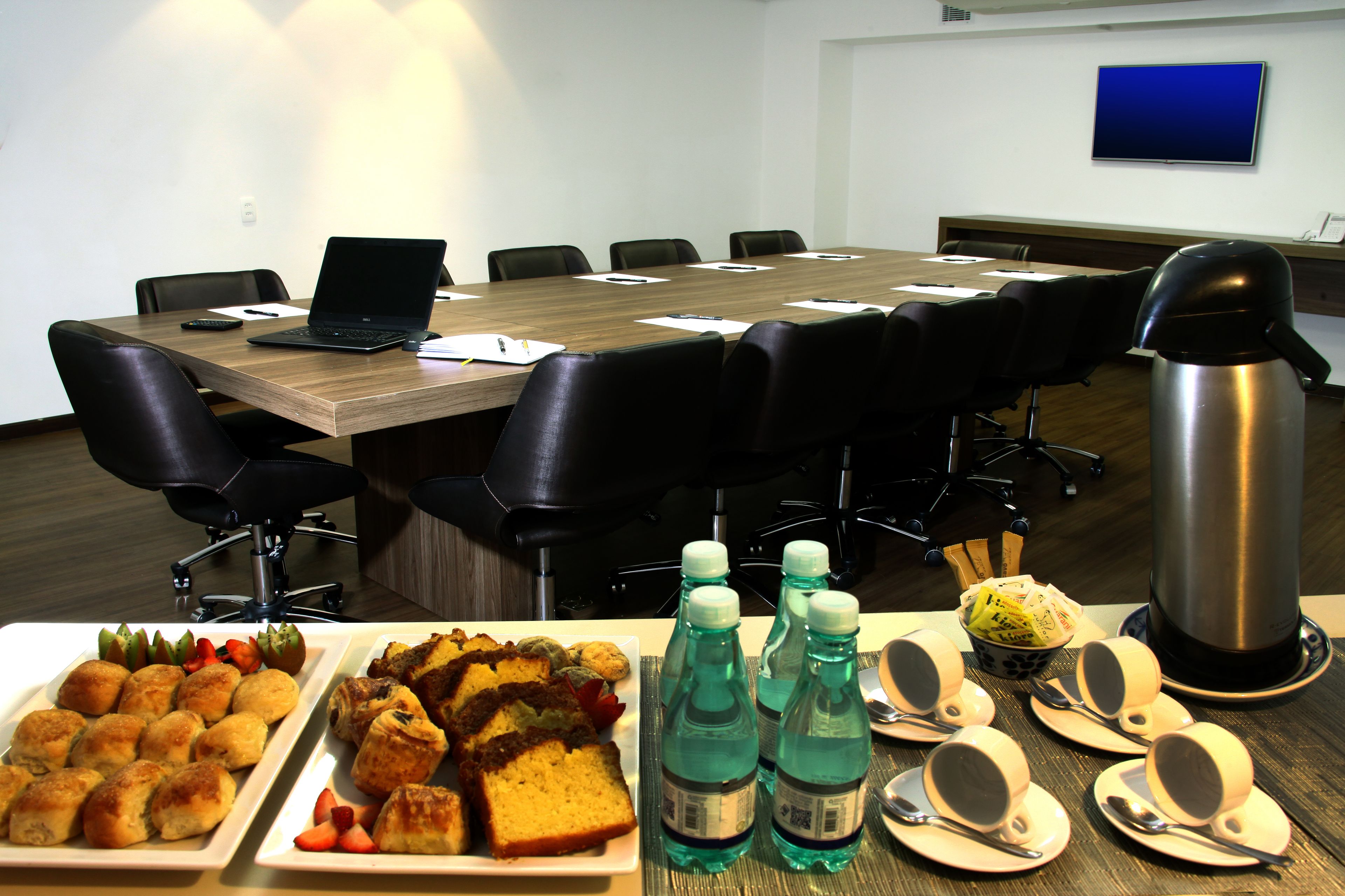 Uma sala de conferência com uma mesa comprida, cadeiras pretas, um laptop e uma cafeteira.