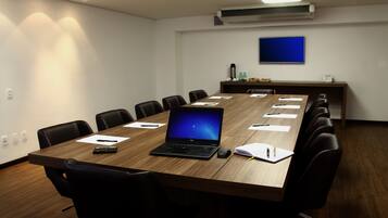 Uma sala de conferências com uma longa mesa de madeira, cadeiras pretas, um laptop, papéis, uma caneta, um telefone e um monitor de parede com tela azul.