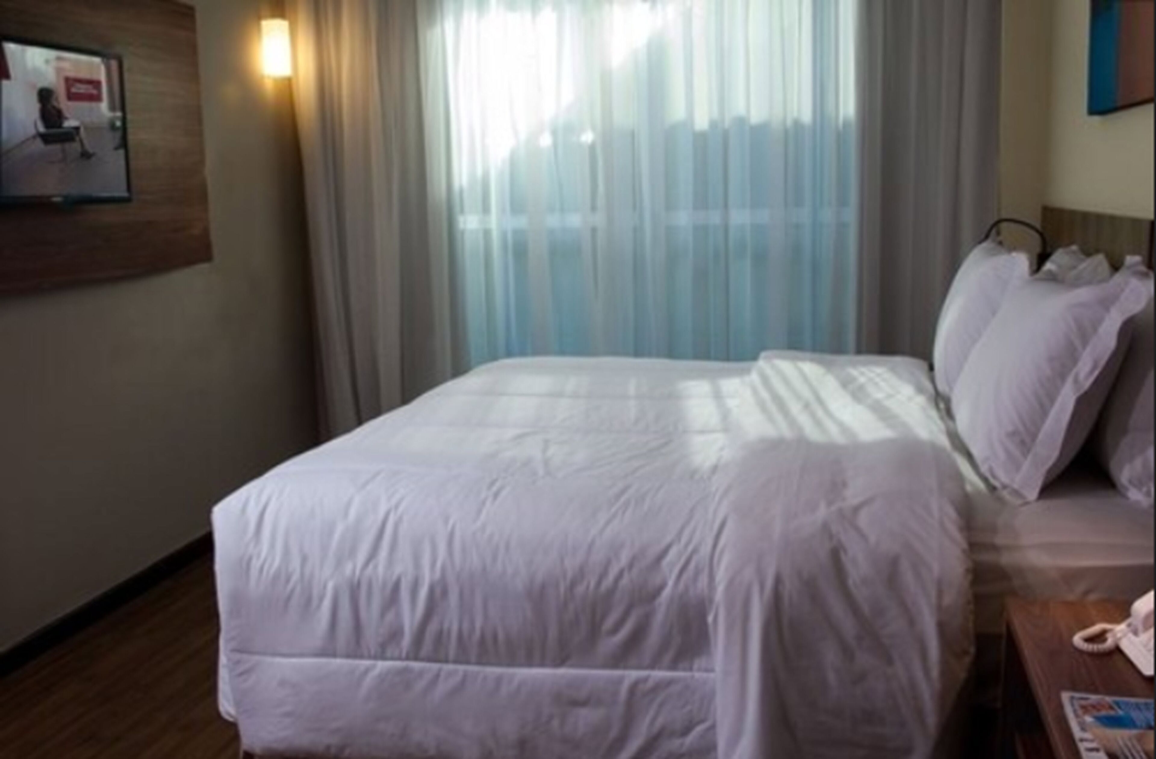 Quarto de hotel com uma cama, uma mesa de cabeceira com telefone e uma janela com cortinas finas.