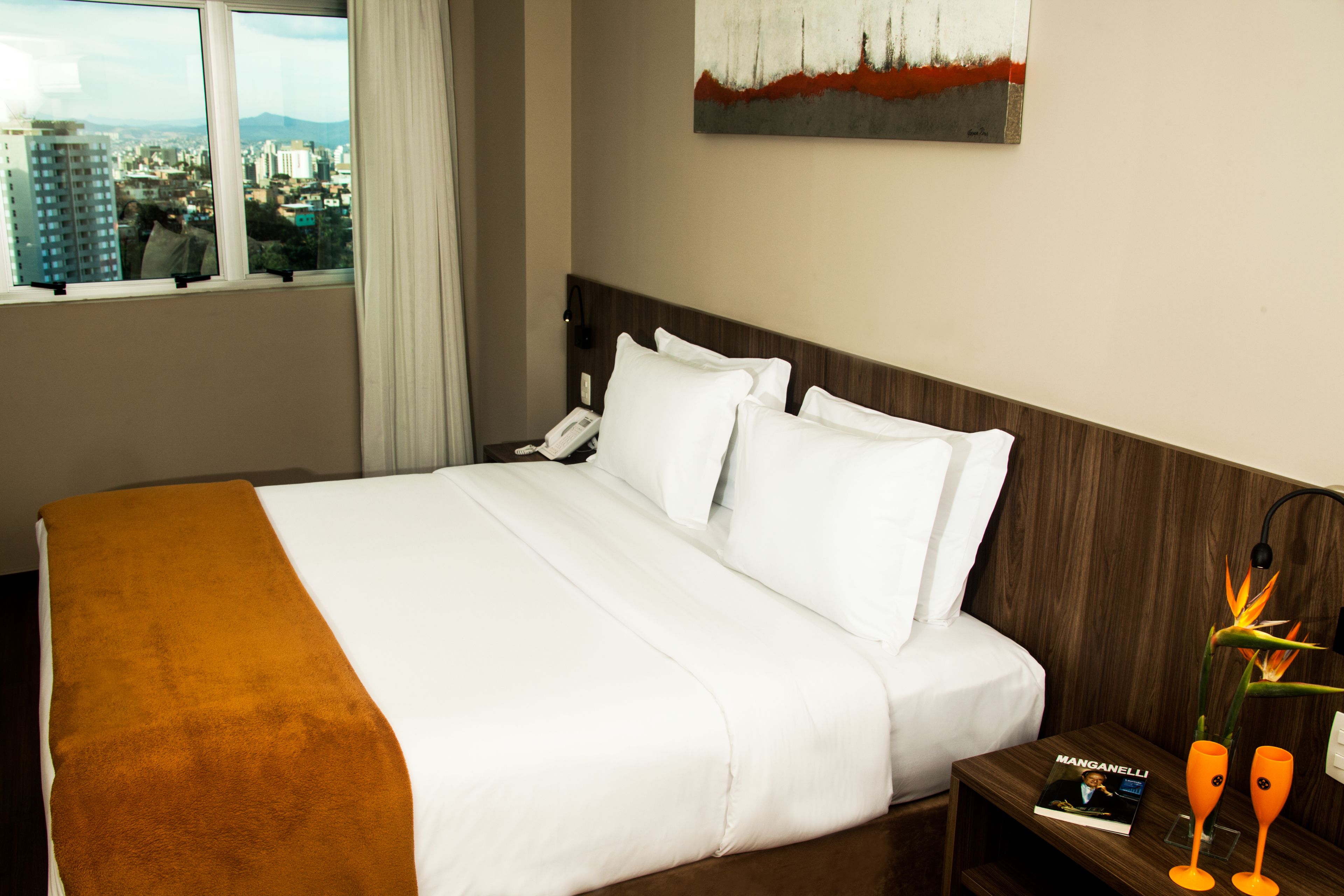 Quarto de hotel com cama, mesa de cabeceira, telefone, revista e janela com vista para a cidade.
