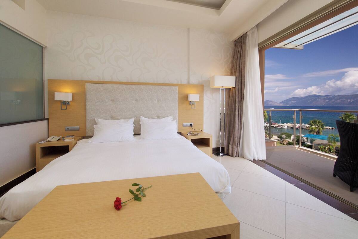 Quarto de hotel com cama, mesas de cabeceira, um criado-mudo, uma pequena mesa com uma rosa e varanda com vista para o mar e barcos.
