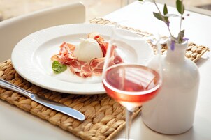 Um prato branco com presunto, mussarela e manjericão sobre um descanso trançado. Também se vê um copo de vinho rosé e um vaso branco com flores roxas.
