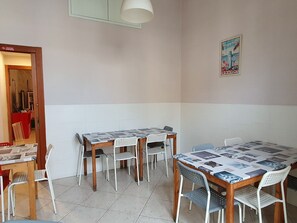 Uma sala de jantar com uma mesa de madeira e cadeiras brancas, piso ladrilhado e luminária fixada na parede.