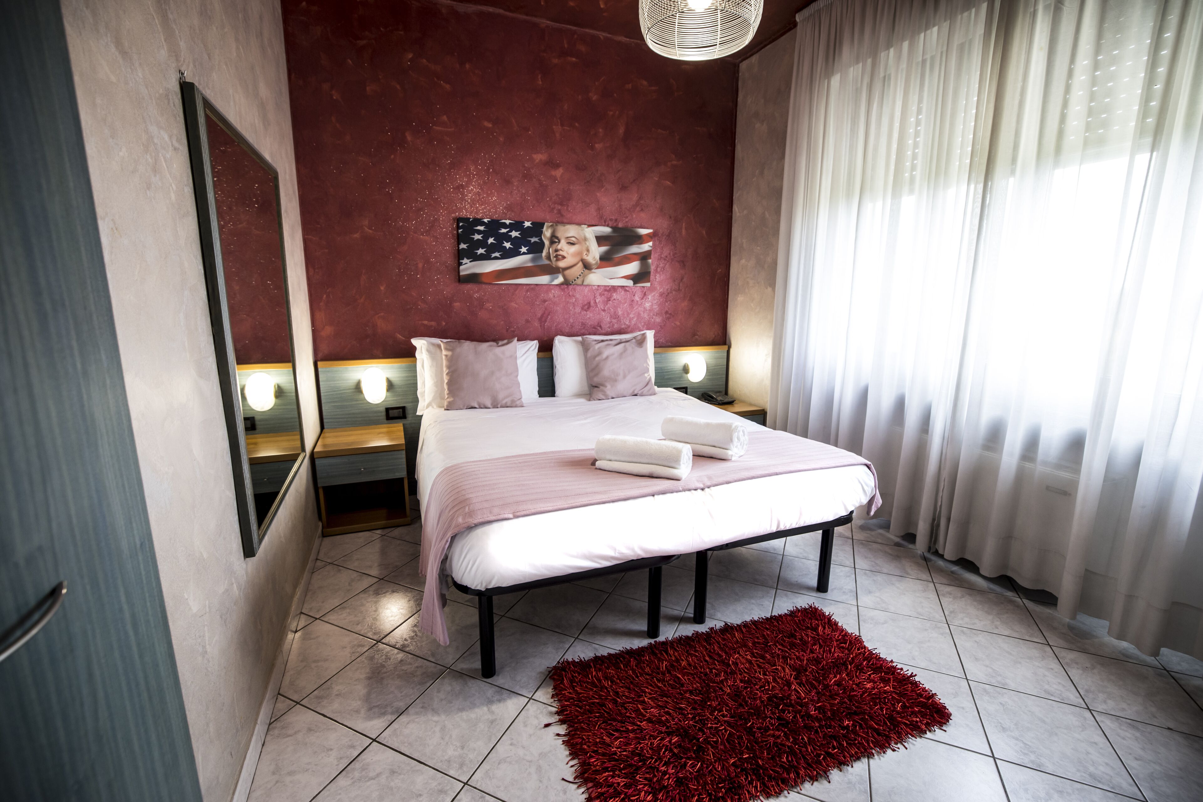 Quarto com cama, tapete vermelho, pintura da bandeira americana e janela com cortinas.