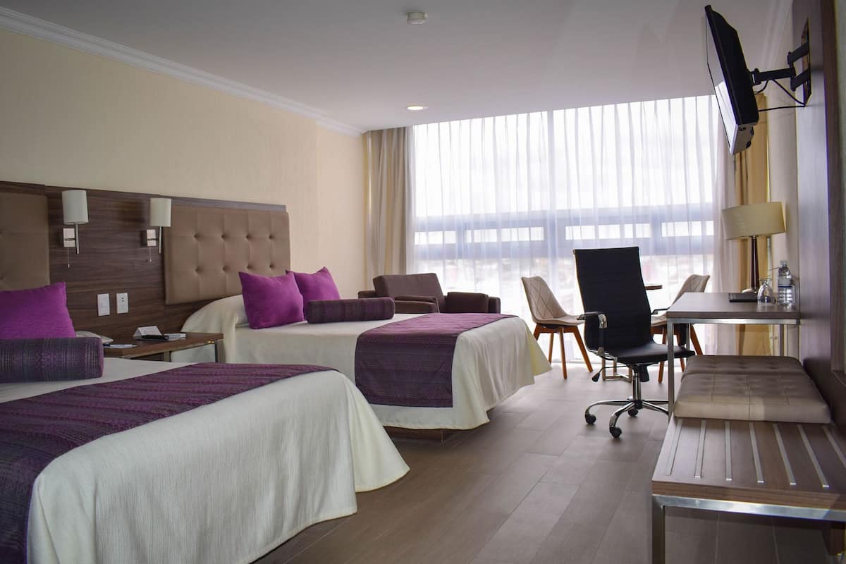 Quarto de hotel com duas camas, uma escrivaninha, uma cadeira, uma televisão e janela com cortinas.