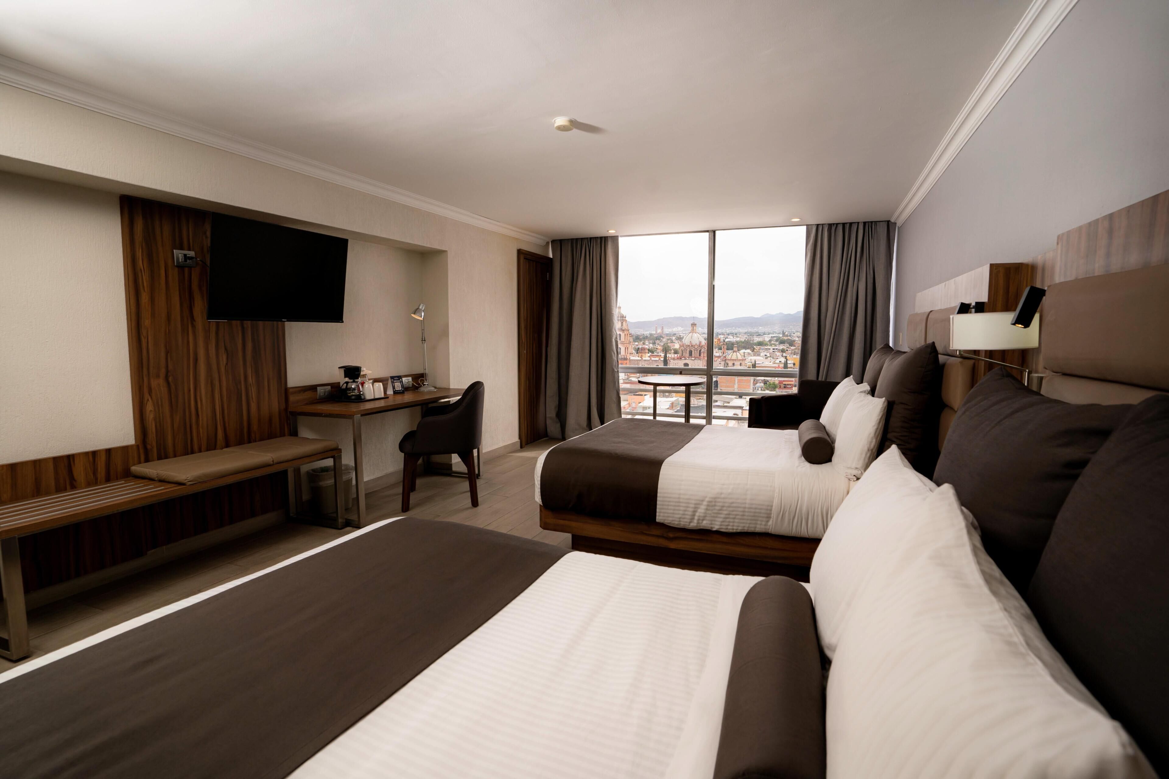 Quarto de hotel com duas camas, uma escrivaninha, uma cadeira, televisão e uma janela grande com vista para a cidade.