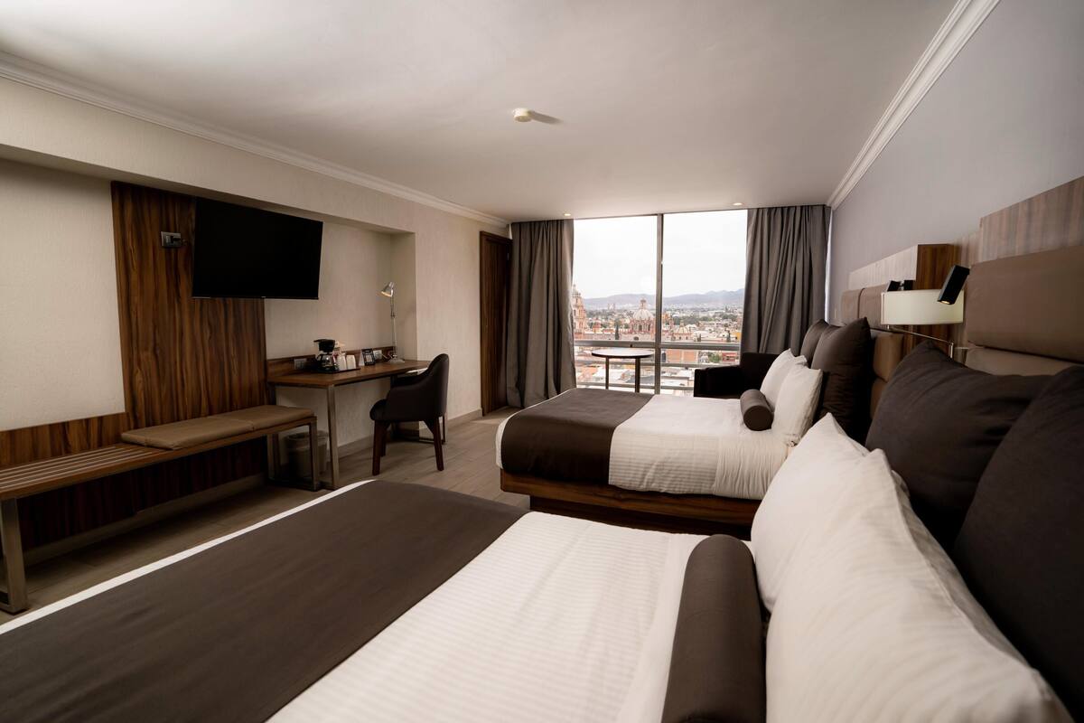 Quarto de hotel com duas camas, uma escrivaninha, uma cadeira, televisão e uma janela grande com vista para a cidade.