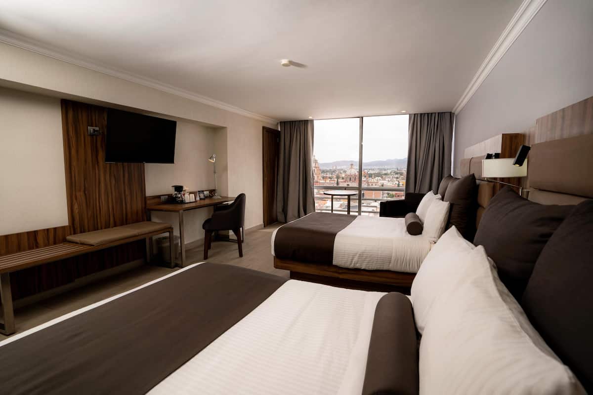 Quarto de hotel com duas camas, uma escrivaninha, uma cadeira, televisão e uma janela grande com vista para a cidade.