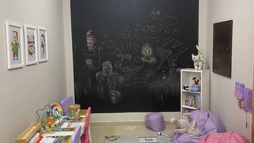 Uma sala de brinquedos infantil com um quadro negro, uma mesa com materiais educativos e um puff.
