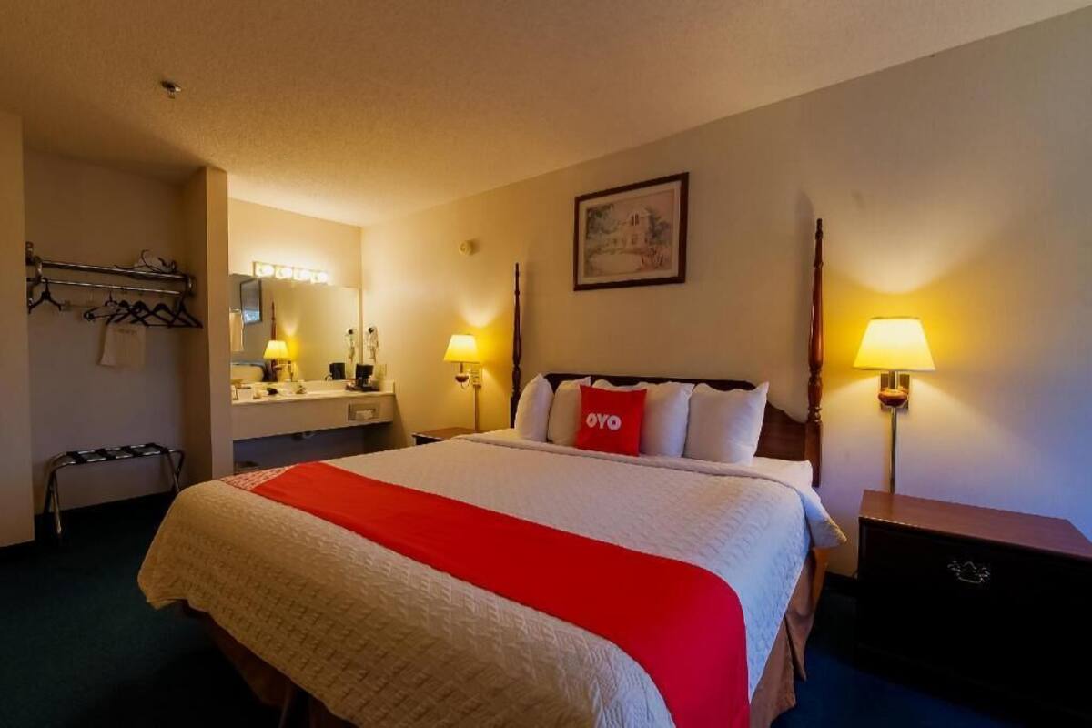 Quarto de hotel com cama, duas luminárias de cabeceira, um espelho, um cabideiro e uma pequena mesa de cabeceira de madeira.