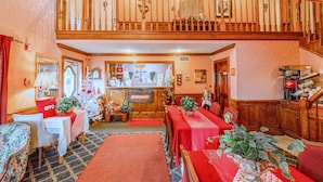 Um saguão de hotel com escadaria de madeira, balcão de recepção e área de refeições com toalhas de mesa vermelhas.