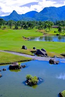Um campo de golfe com um lago, palmeiras e montanhas ao fundo.
