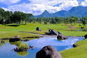Um campo de golfe com um lago, palmeiras e montanhas ao fundo.