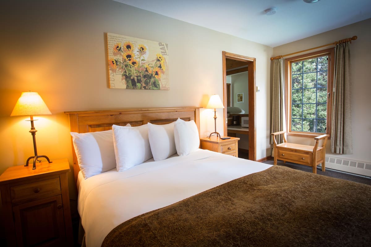 Quarto de hotel com uma cama grande, duas mesas de cabeceira com luminárias, uma cadeira de madeira pequena e uma janela com cortinas.