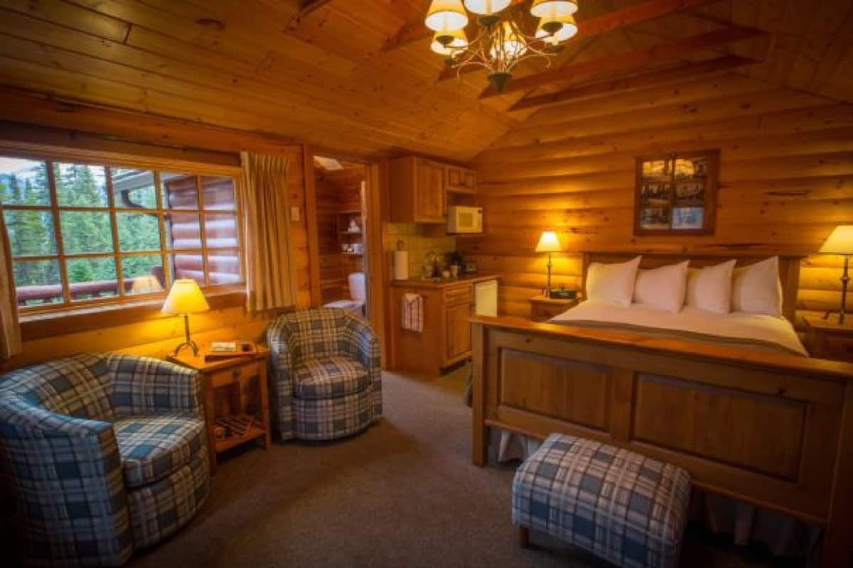 Quarto acolhedor de cabana de madeira com cama, duas poltronas xadrez, uma mesinha e uma janela com cortinas.