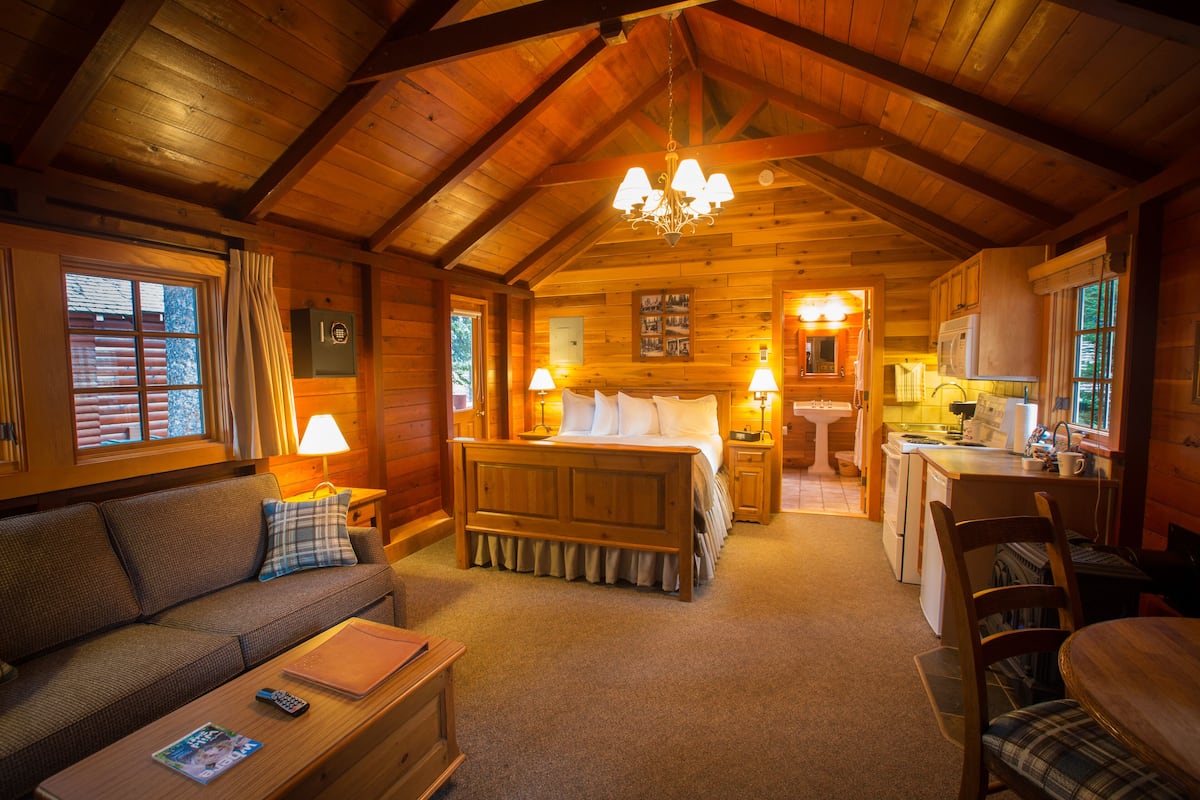 Interior acolhedor de uma cabana de madeira, com uma cama de madeira, um sofá, uma mesa de jantar e uma área de cozinha.