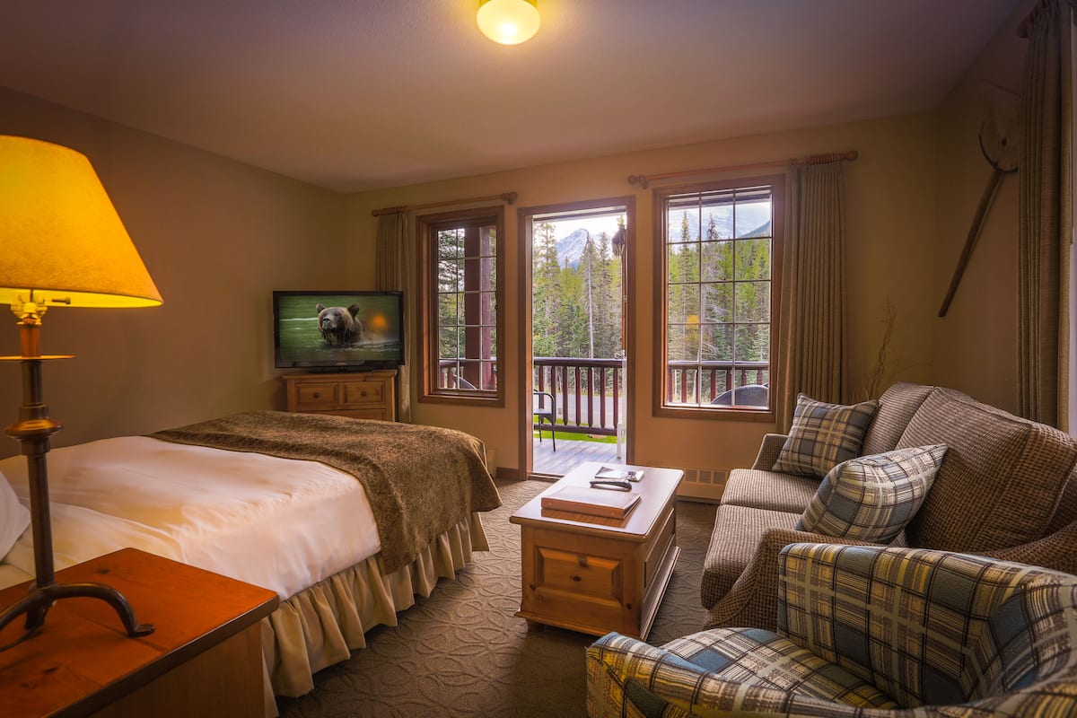 Quarto de hotel com cama, sofá, televisão e varanda com vista para árvores e montanhas.
