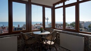 Quarto com janelas amplas, cadeiras de vime, uma mesa pequena e vista para o mar e a paisagem urbana.