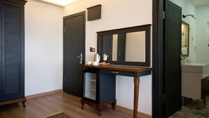 Quarto com piso de madeira, escrivaninha escura com espelho, um pequeno armário e porta para o banheiro.