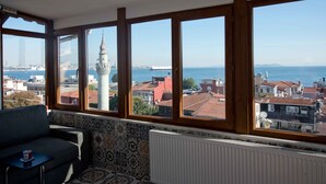 Quarto com grandes janelas que oferecem vista para a paisagem urbana, incluindo uma torre imponente e o mar.