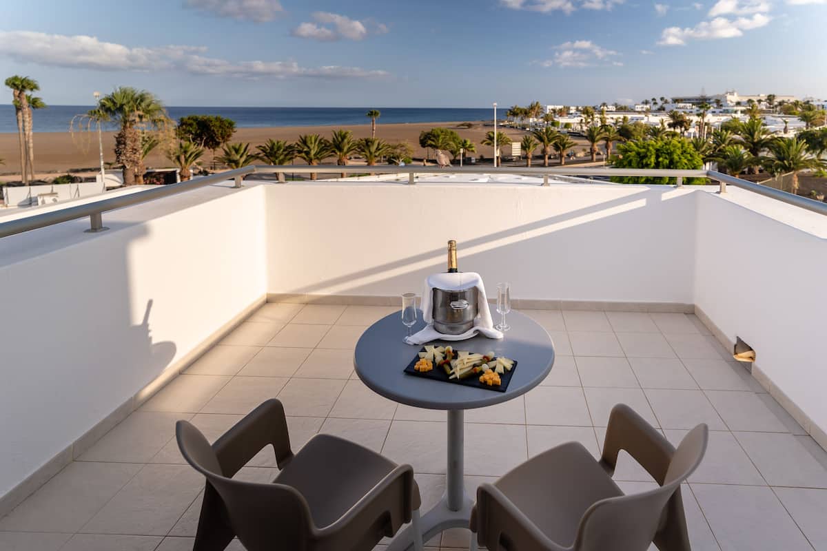 Terraço com mesa posta para dois, uma garrafa de champanhe e um prato de petiscos. O terraço tem vista para uma praia e palmeiras.