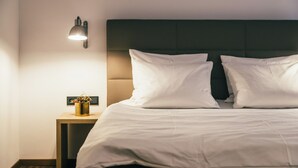 Cama bem arrumada com roupa de cama branca e cabeceira escura. Tem uma mesinha de cabeceira pequena com uma luminária e uma planta.