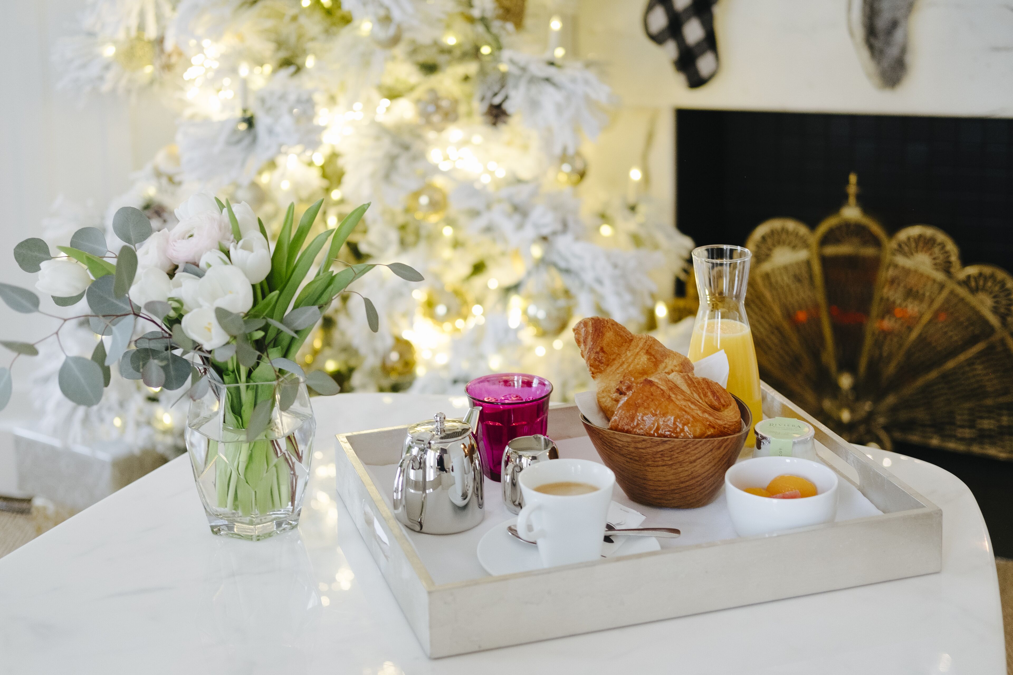 Uma bandeja de café da manhã com croissant, suco de laranja, café e um vaso de flores sobre uma mesa de mármore, com um pinheiro de Natal branco ao fundo.