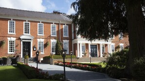Um edifício de tijolos com entrada central, degraus que a conduzem e um jardim com flores e uma árvore.