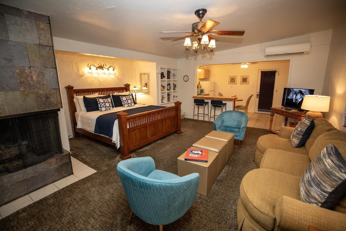 Quarto de hotel com cama, lareira, sofá, poltrona, TV e área de cozinha ao fundo.