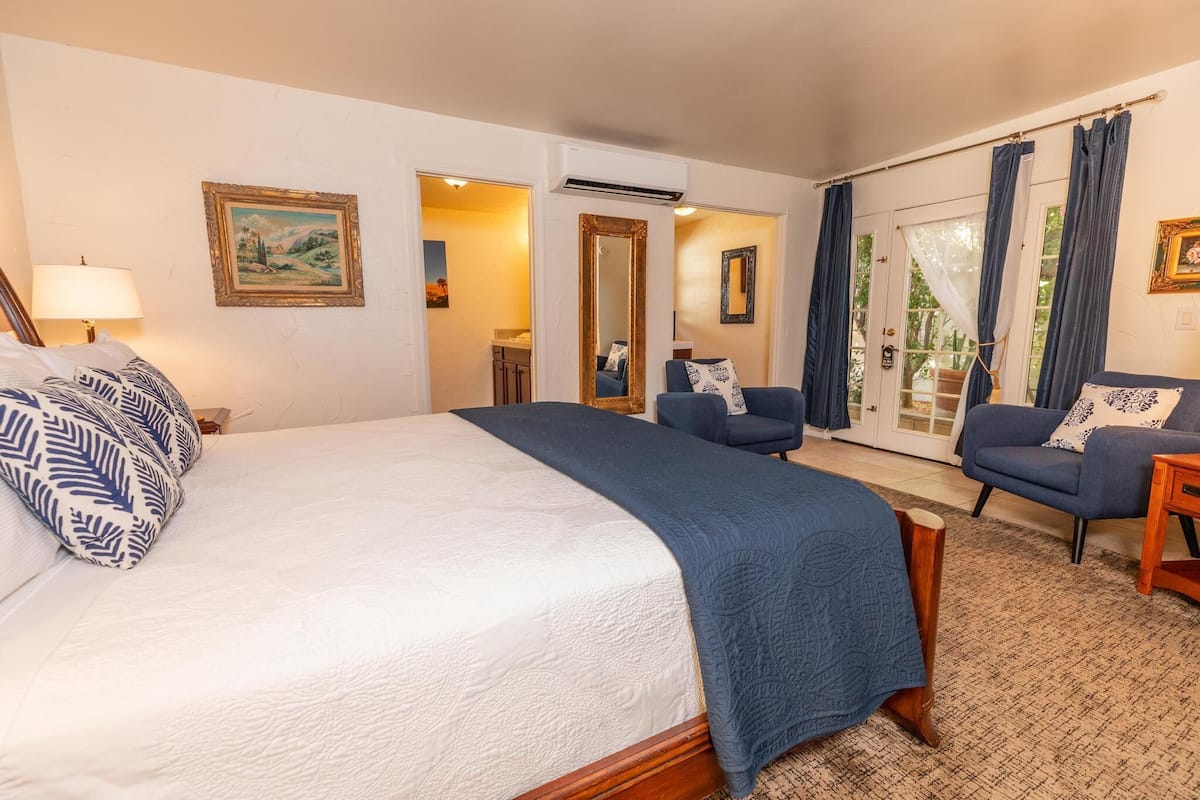 Quarto de hotel com uma cama grande, uma escrivaninha, uma cadeira, um sofá e vista para o exterior através de uma porta de vidro.