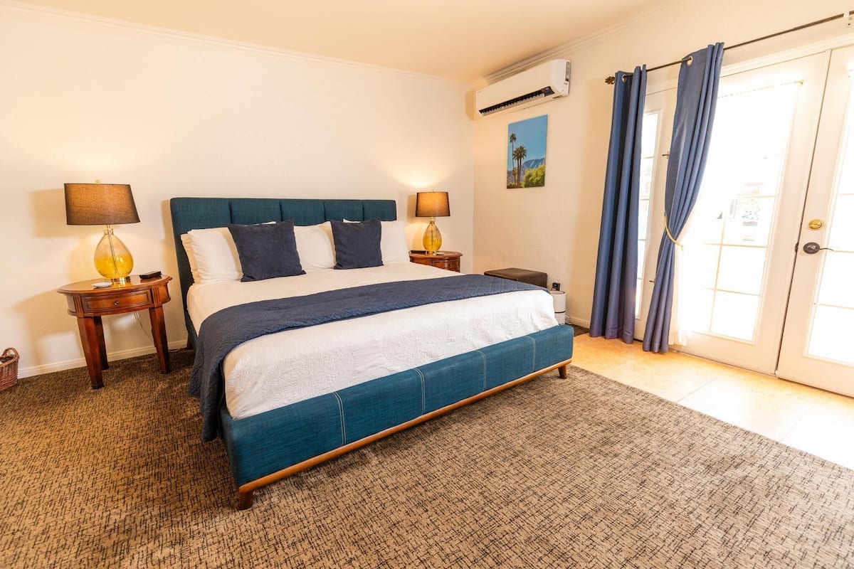 Quarto com cama, duas mesas de cabeceira, luminária, ar-condicionado fixado na parede e vista através de uma porta.