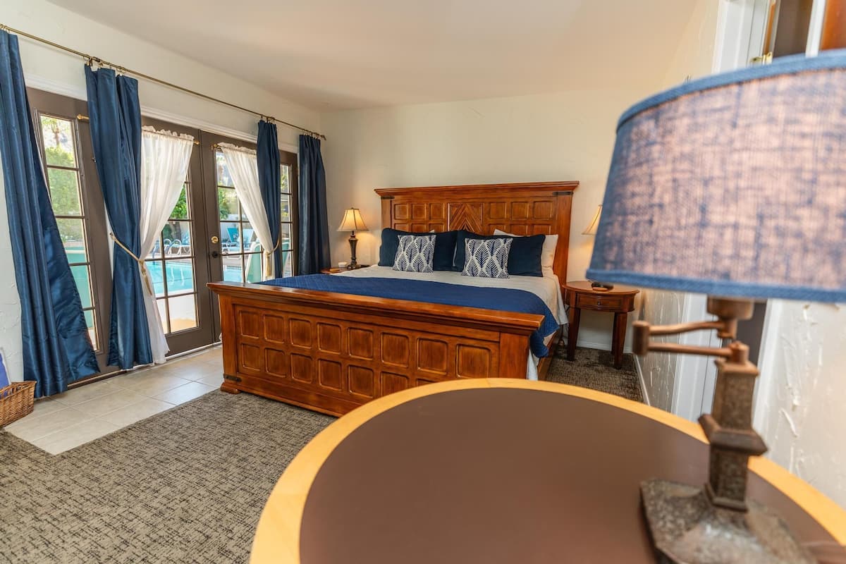 Quarto com cama de madeira, mesa de cabeceira, luminária e vista para uma piscina através das portas de vidro.