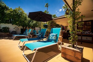 Área ao redor da piscina com espreguiçadeiras e um guarda-sol, cercada por vegetação e um edifício com uma placa visível.