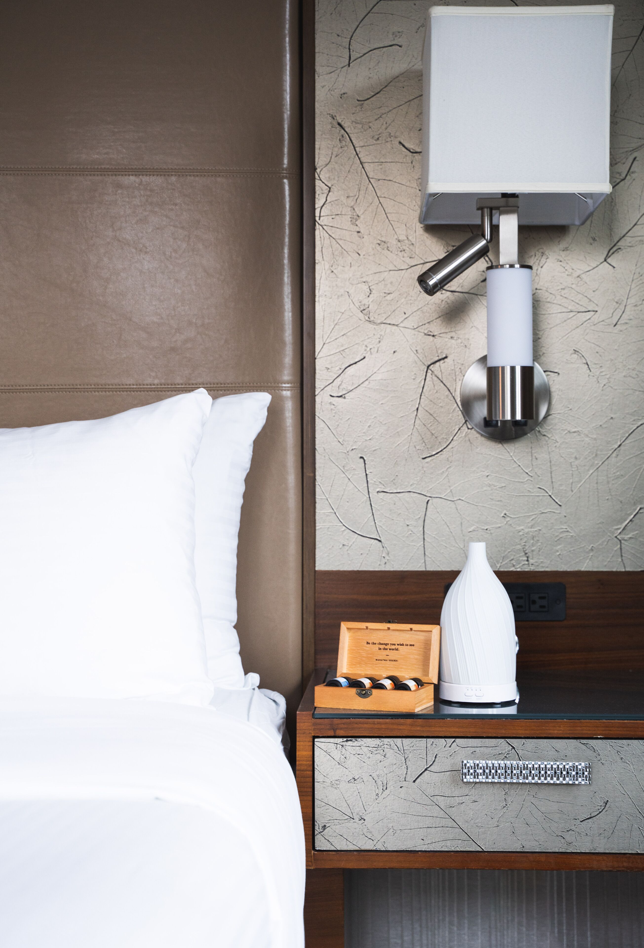 Cama bem arrumada com lençóis brancos, mesa de cabeceira com um vaso branco, caixa de madeira e luminária fixada na parede.
