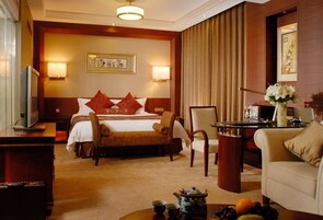 Quarto de hotel com cama, escrivaninha, cadeira, televisão e mesa de centro com bule e frutas.