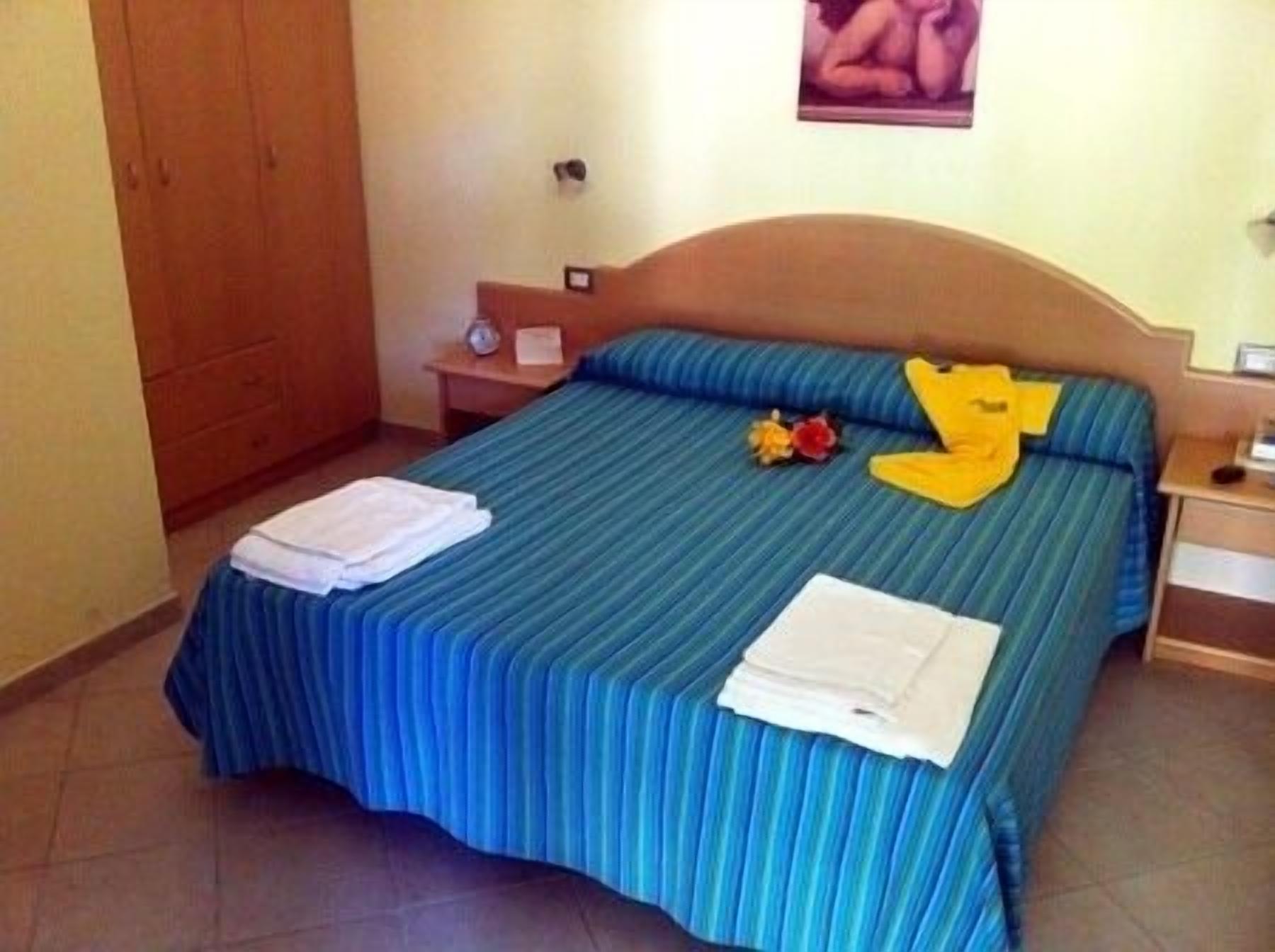 Quarto de hotel com cama, mesa de cabeceira, relógio de parede e uma moldura com imagem.