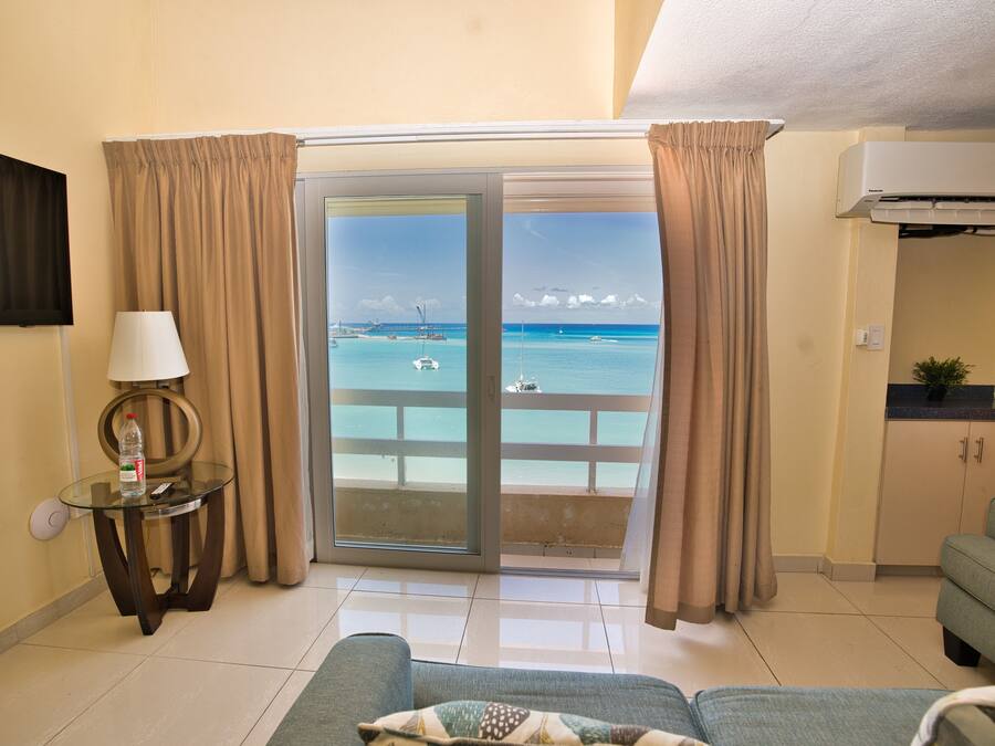 Quarto com sofá, mesa de vidro com uma garrafa, cortinas, TV e vista para o oceano.