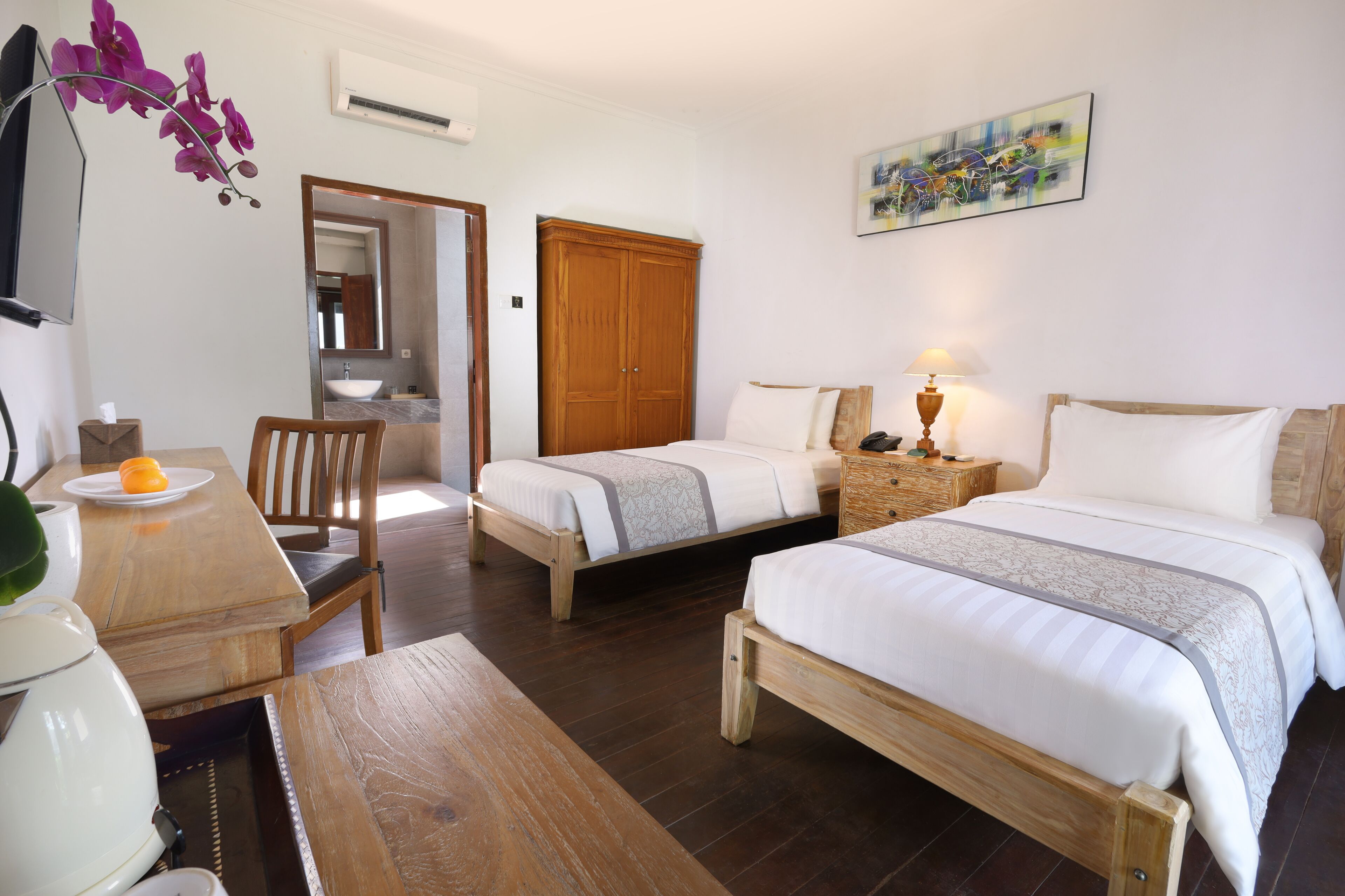 Quarto de hotel com duas camas de solteiro, uma mesa de madeira com uma cadeira, uma pequena mesa de cabeceira com uma luminária e um quadro na parede.