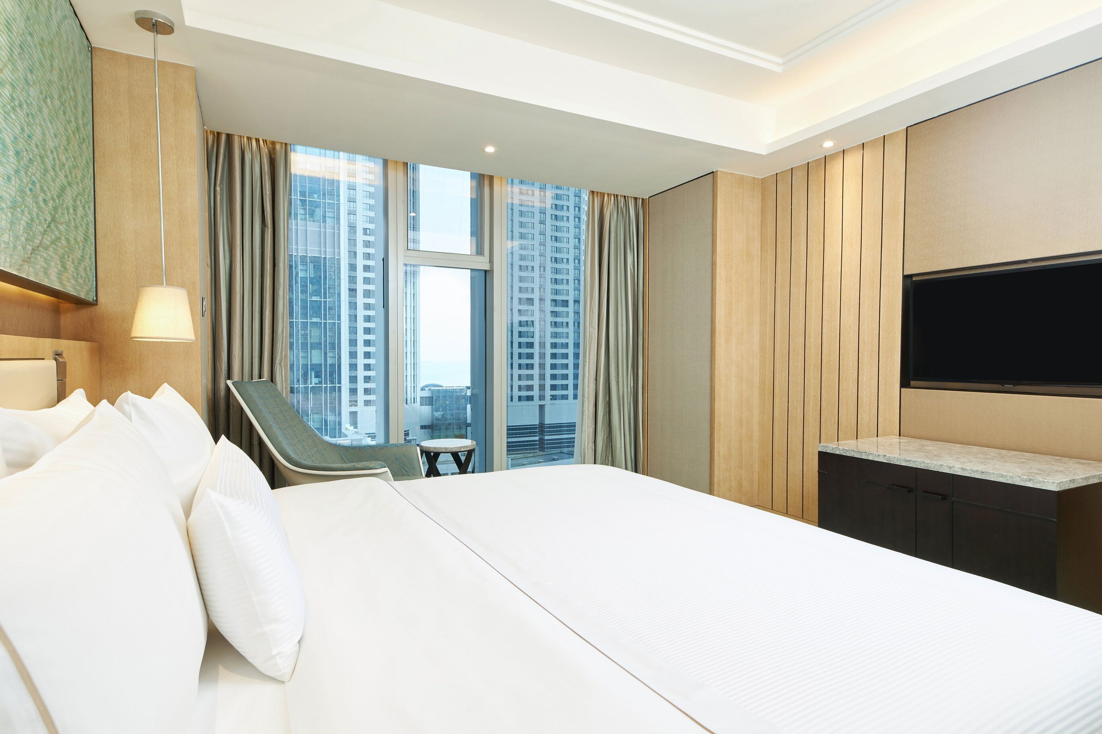 Quarto de hotel com uma cama grande, televisão na parede, escrivaninha com cadeira e vista para edifícios altos pela janela.