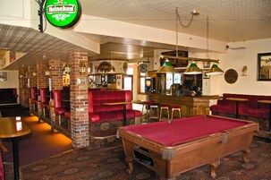 Uma mesa de bilhar, assentos com almofadas vermelhas e um bar com um anúncio da Heineken.