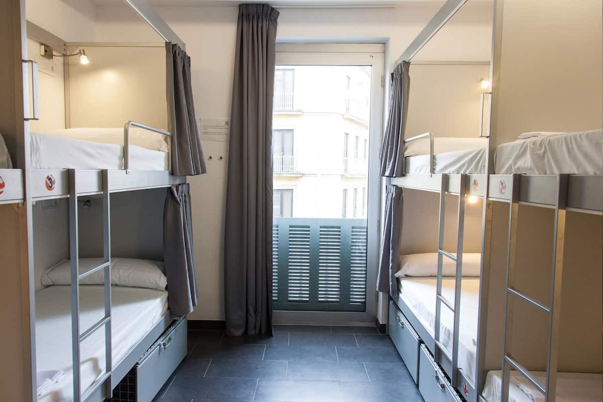 Quarto de hotel compacto com beliches, uma janela com persianas e vista para edifícios.