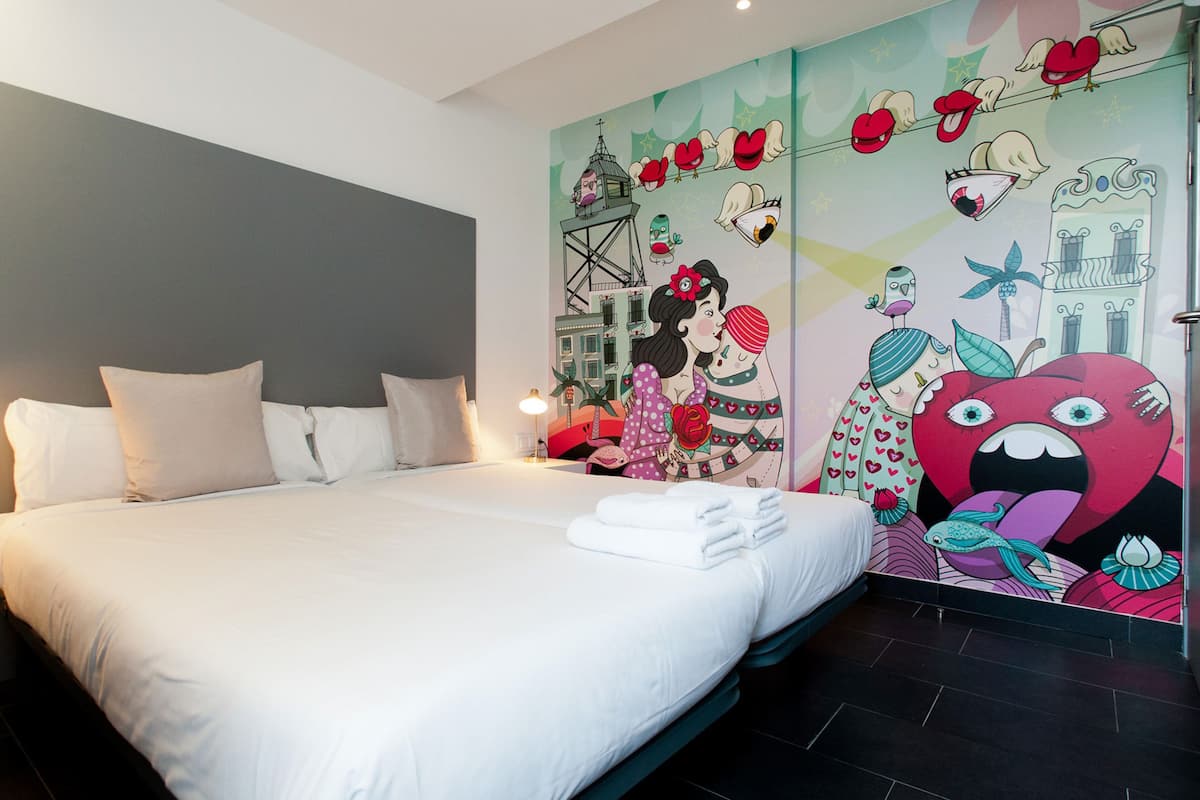 Quarto de hotel com uma cama grande, roupa de cama branca e um mural colorido na parede.
