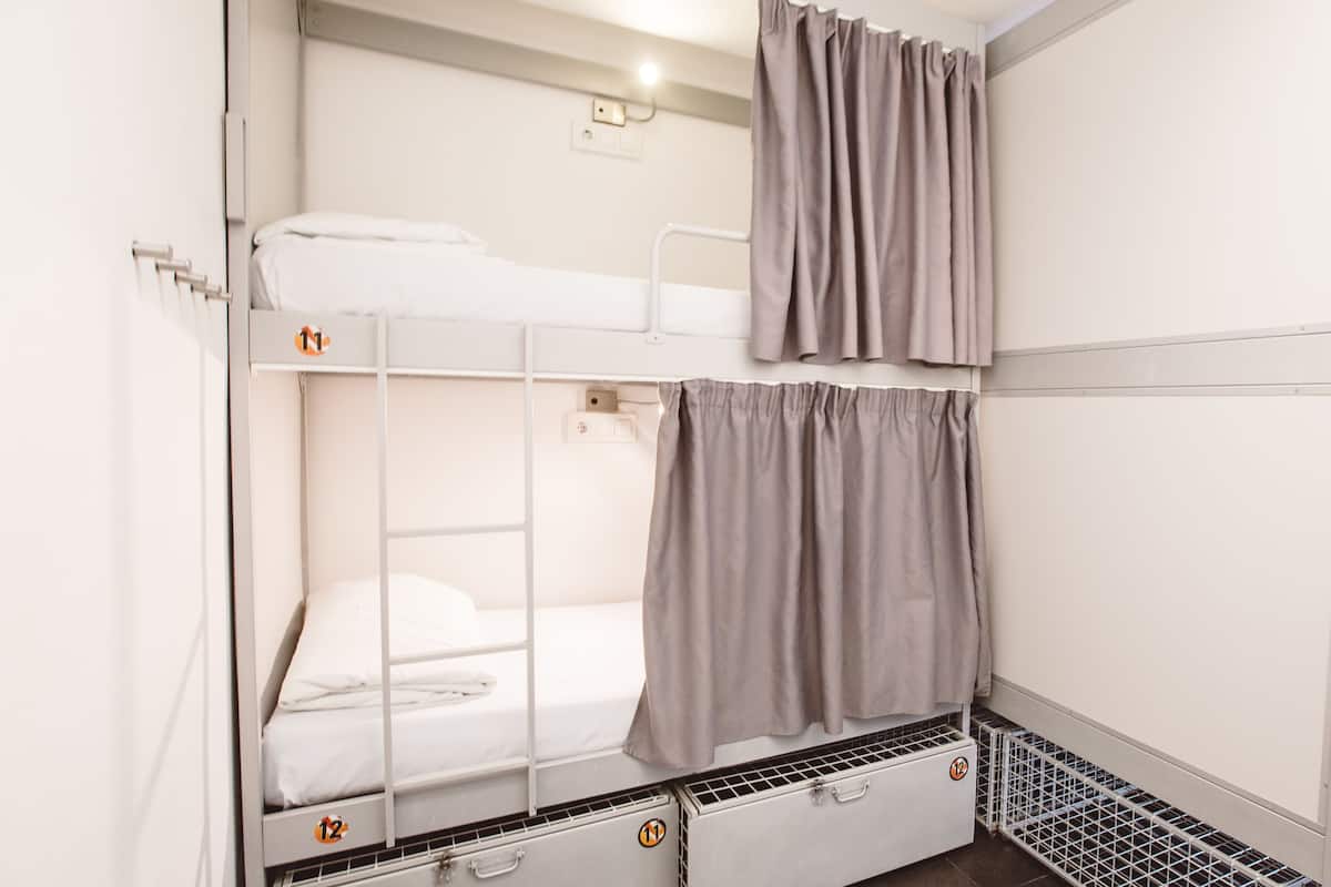 Quarto com beliche, uma escrivaninha pequena e uma janela com cortinas.