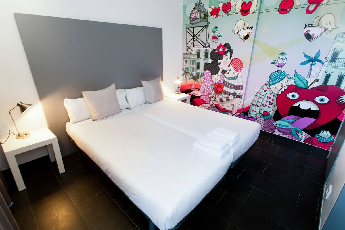 Quarto de hotel com uma cama grande, roupa de cama branca, mesa de cabeceira com abajur e um mural colorido na parede.